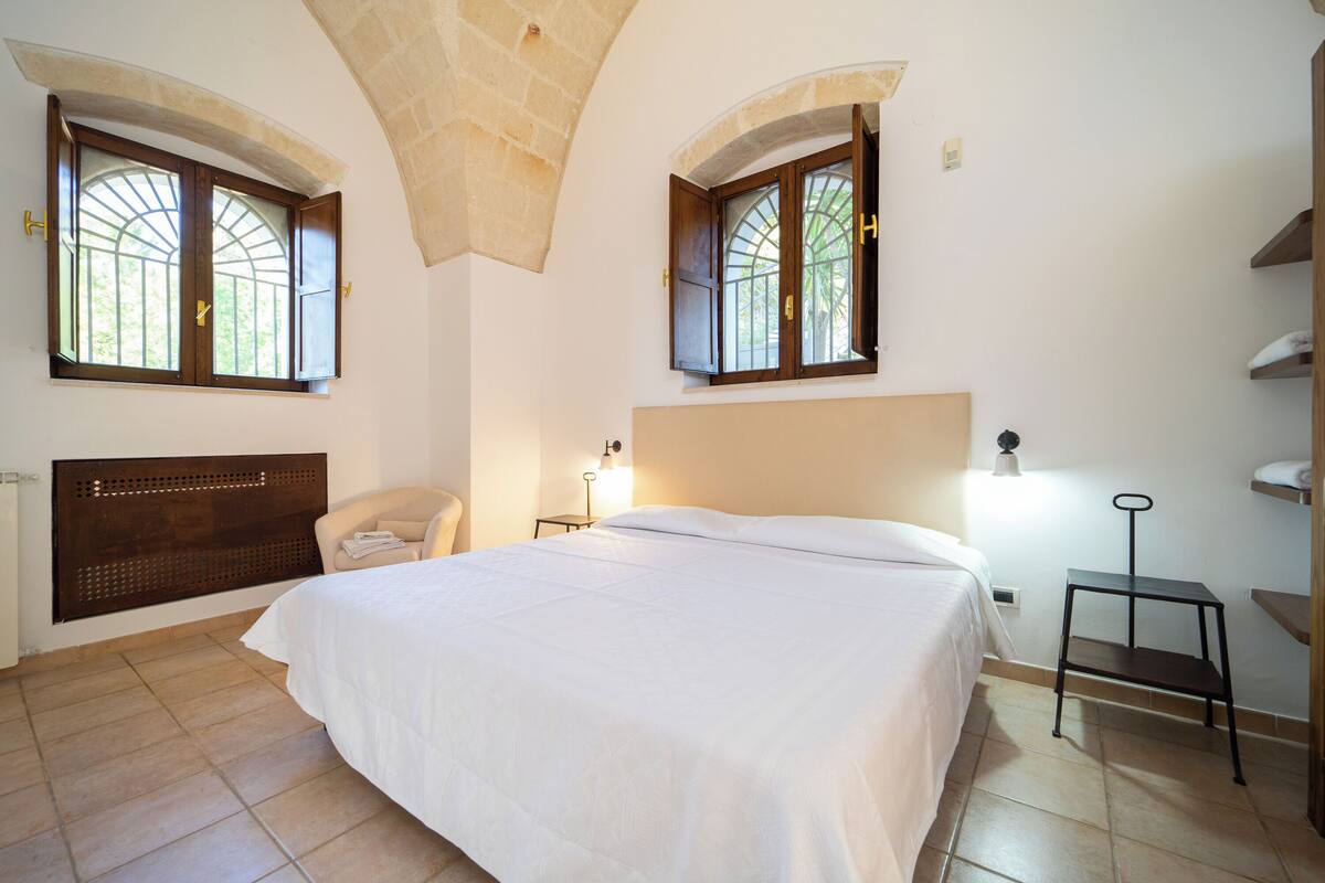 Quarto com cama grande, duas janelas com molduras de madeira, mesa de cabeceira com abajur e uma cadeira.