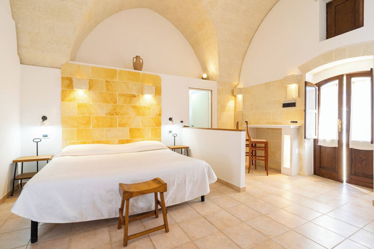 Quarto de hotel com uma cama grande, um banquinho de madeira, uma mesa pequena e uma parede de pedra.