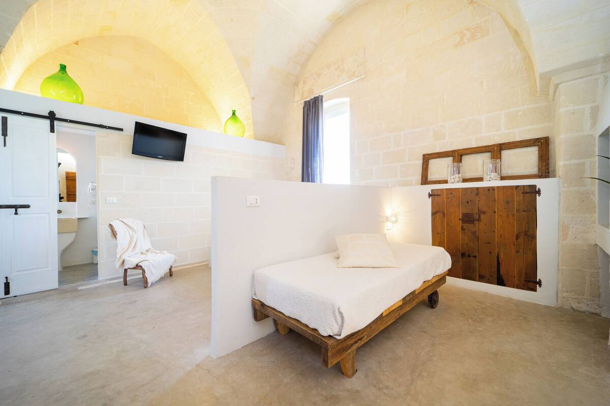 Quarto com cama de madeira, uma cadeira branca, televisão fixada na parede e porta de madeira com painéis de vidro.