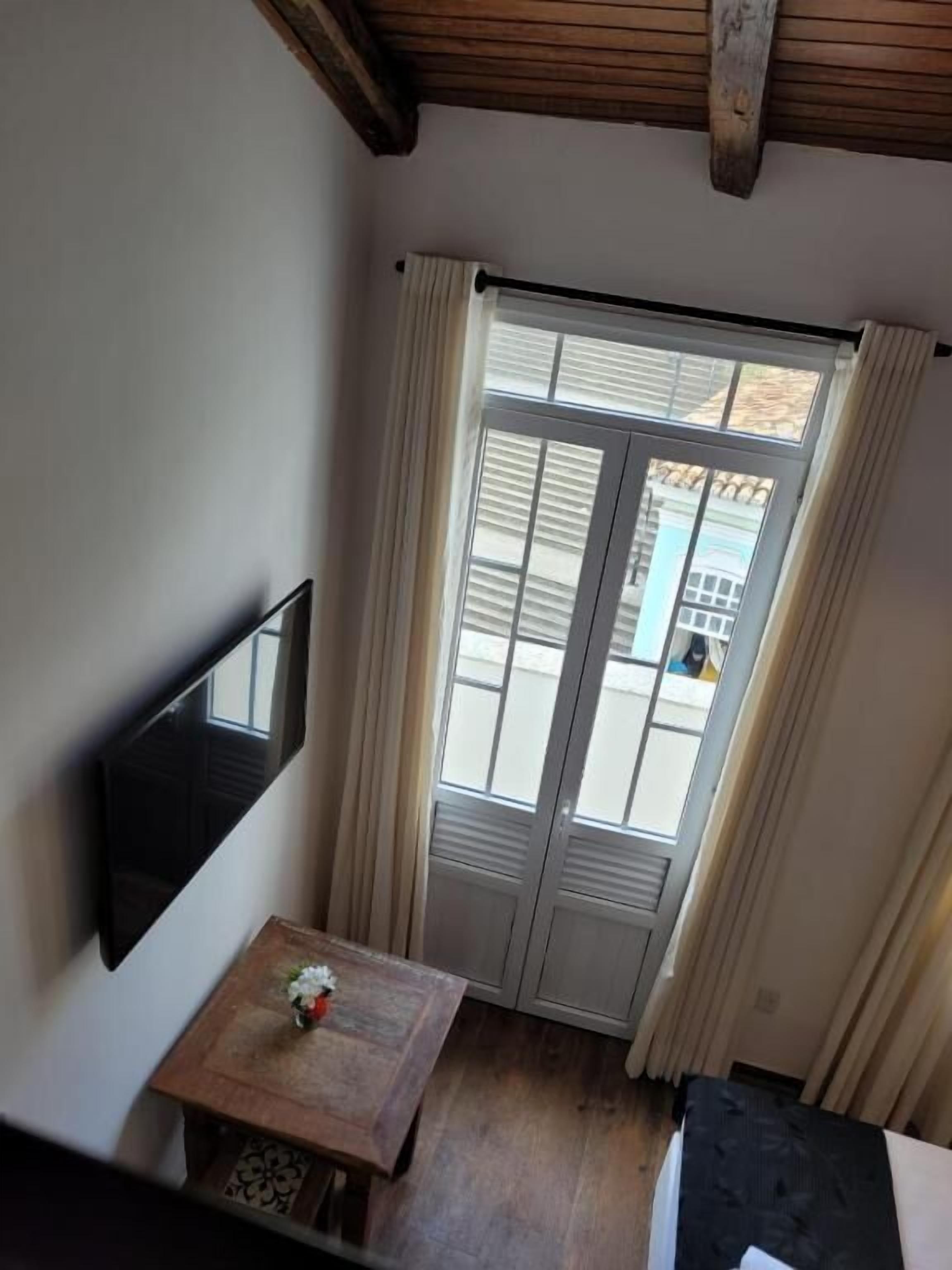 Quarto com cama, uma pequena mesa com uma flor, televisão na parede e porta de vidro deslizante para o exterior.