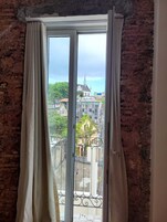 Quarto com uma janela ampla, vista para uma igreja e varanda com grade de metal.