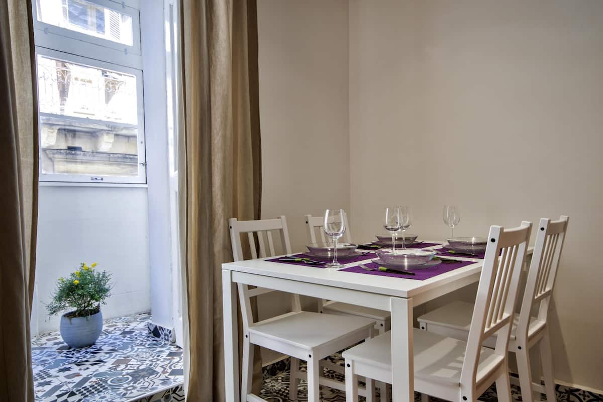 Mesa de jantar posta com cadeiras brancas, um vaso de planta e uma janela com cortinas.