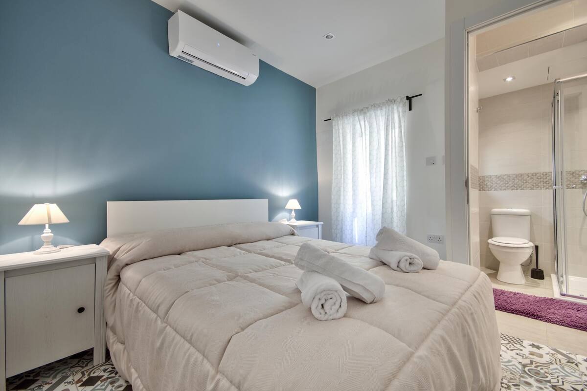 Quarto com cama, duas mesas de cabeceira com luminárias, banheiro com vaso sanitário e chuveiro, e uma parede azul.