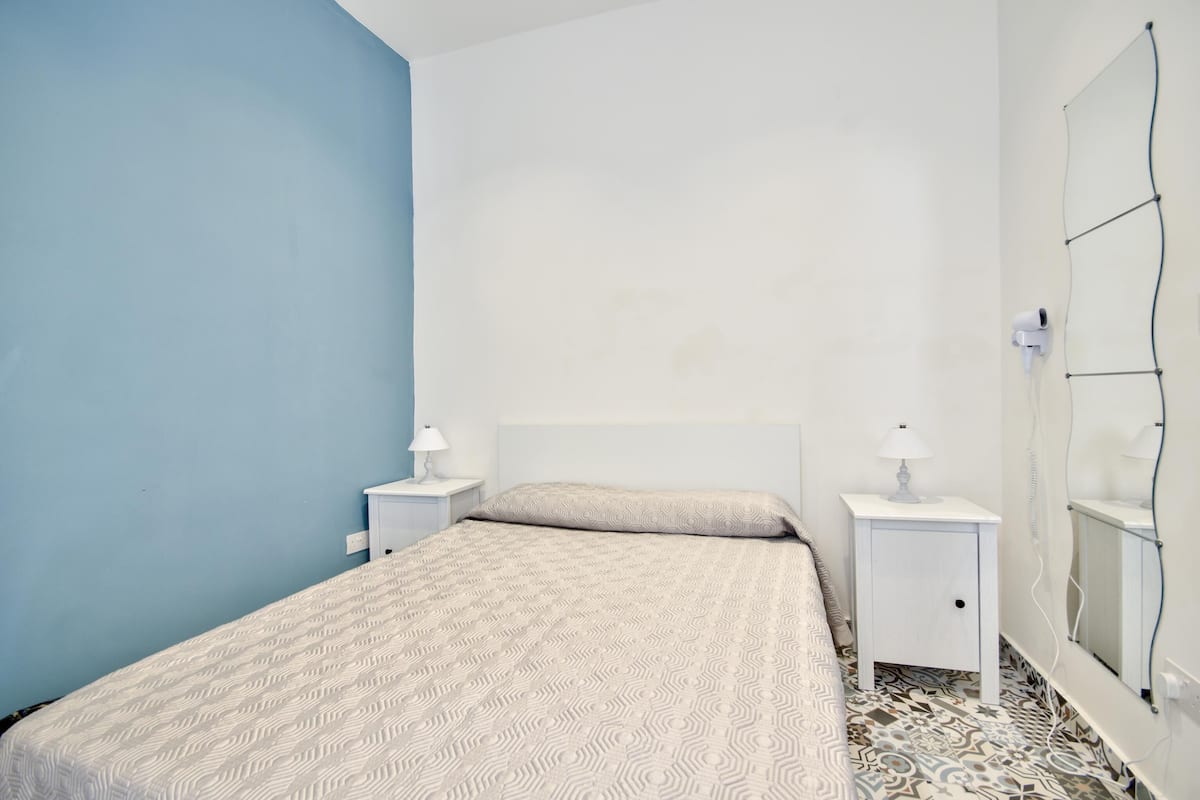 Quarto com cama, duas mesas de cabeceira com abajures, um espelho fixado na parede e um piso estampado.
