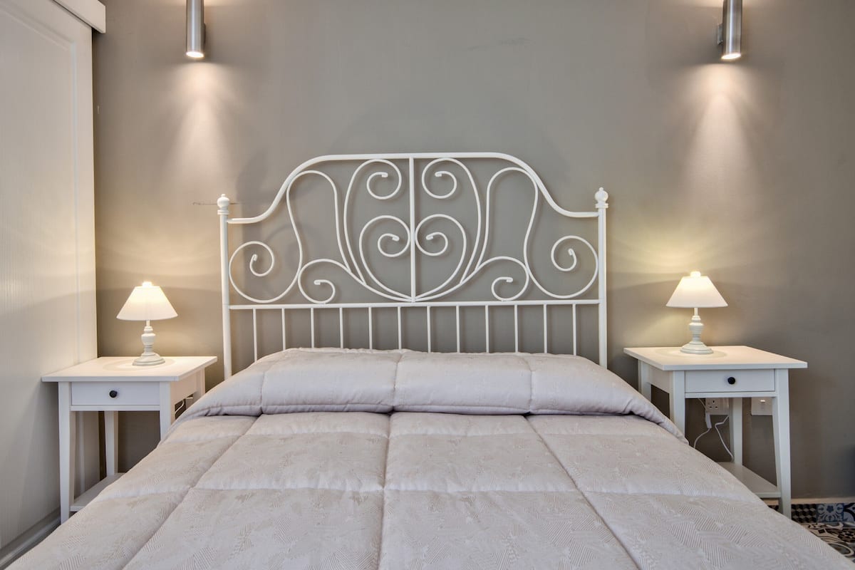 Cama com cabeceira de metal, duas mesas de cabeceira com luminárias e parede em tom neutro.