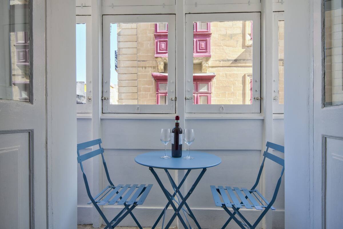 Um quarto com uma mesa pequena, duas cadeiras, uma garrafa de vinho e dois copos de vinho.
