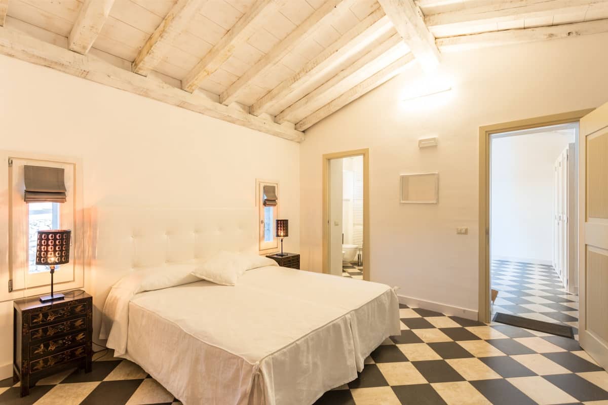 Quarto com piso xadrez, cama com roupa de cama branca, mesa de cabeceira com abajur e espelho na parede.