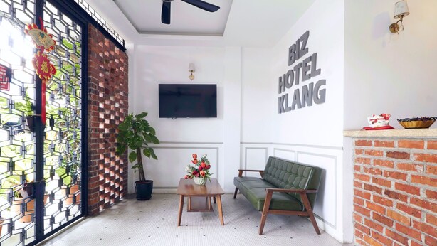 Quarto de hotel com parede de tijolos, um sofá verde, mesa de madeira com um vaso de flores e televisão na parede.