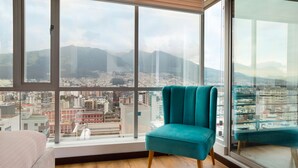 Quarto com uma janela ampla que oferece vista para a cidade, uma cadeira verde-água e uma cama.