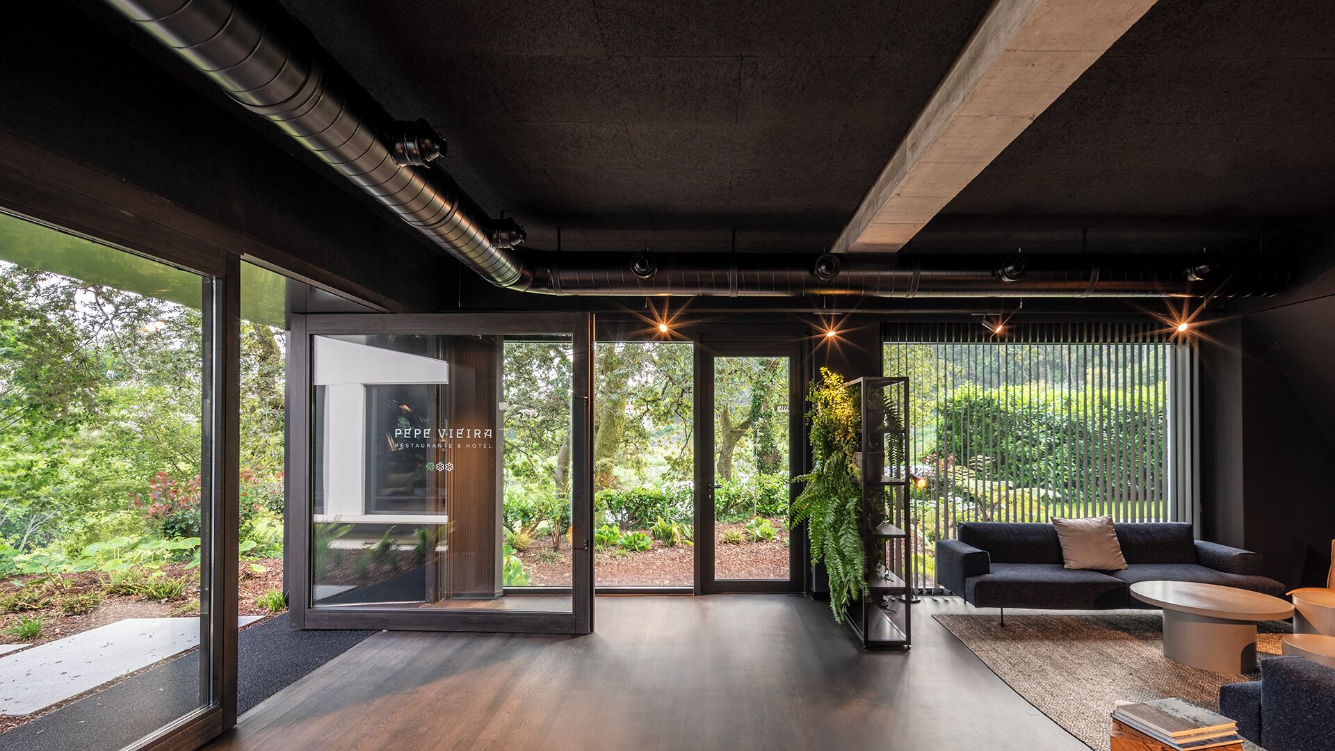 Um interior moderno com grandes portas de vidro que dão para um jardim exuberante, um sofá de cor escura e uma mesa de centro minimalista.