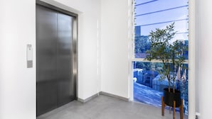 Um elevador moderno com acabamento metálico, uma planta em vaso perto de uma janela ampla e vista desimpedida para o exterior.