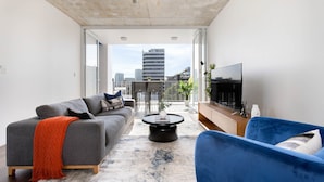 Uma sala de estar moderna com um sofá cinzento, poltrona azul e uma mesa de centro. Uma televisão está instalada na parede, e há uma varanda com vista para a cidade.
