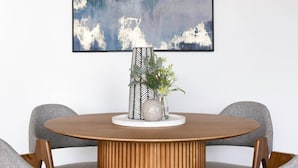 Mesa redonda de madeira com duas cadeiras cinzas estofadas, uma pintura abstrata emoldurada acima e um vaso com folhagem sobre a mesa.