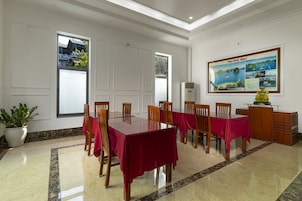 Uma sala de conferências com uma mesa de vidro, toalhas de mesa vermelhas, cadeiras de madeira, uma planta em vaso e um mapa emoldurado da Baía de Halong na parede.