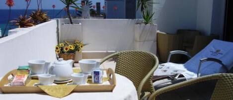 Uma varanda com mesa posta para o café da manhã, cadeiras de vime e vista para o mar e edifícios.