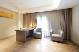 Quarto de hotel moderno com piso de madeira, uma escrivaninha, duas poltronas, uma mesinha e uma grande janela com cortinas.