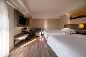 Quarto de hotel com duas camas, uma escrivaninha com cadeira, televisão na parede e abajur na mesinha de cabeceira.