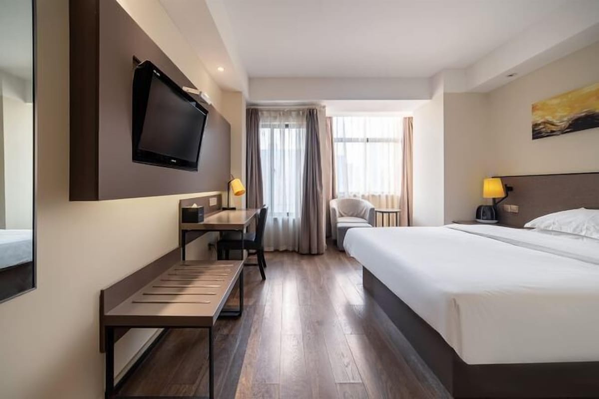 Quarto de hotel com cama, escrivaninha, cadeira, TV fixada na parede e janela com cortinas.