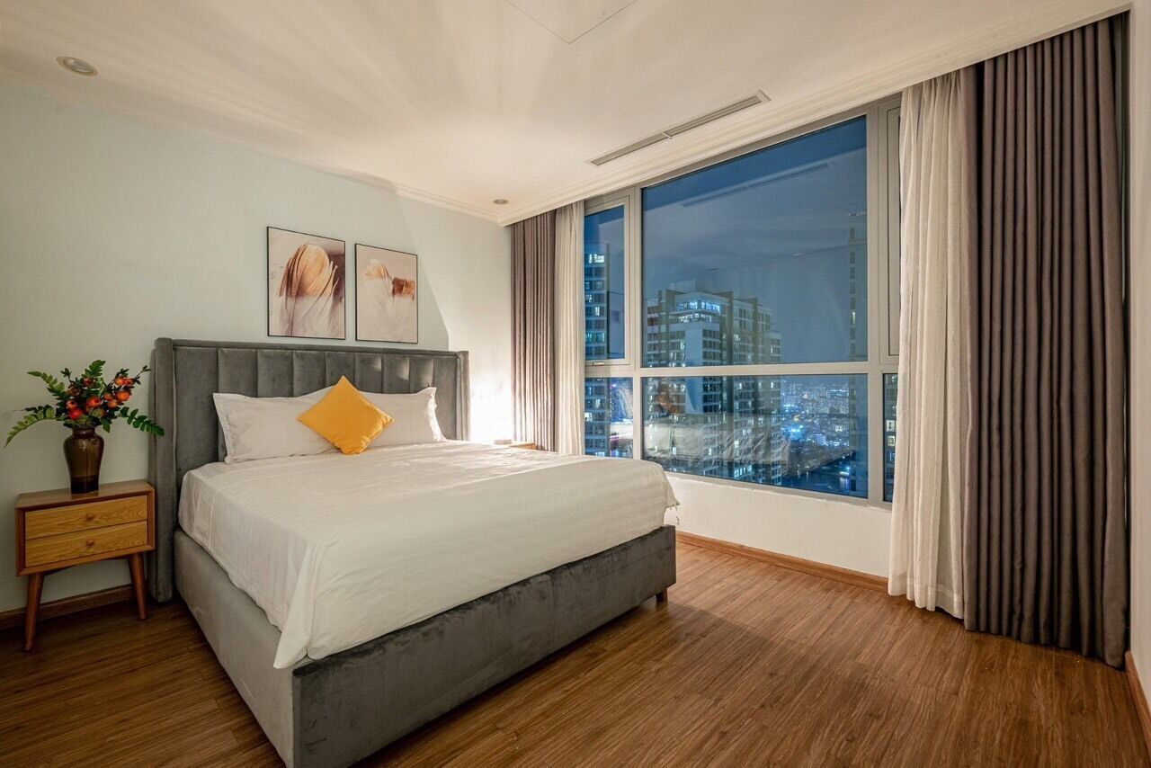 Quarto de hotel moderno com uma cama grande, uma mesa de cabeceira, uma planta em vaso e uma grande janela com vista para a cidade.