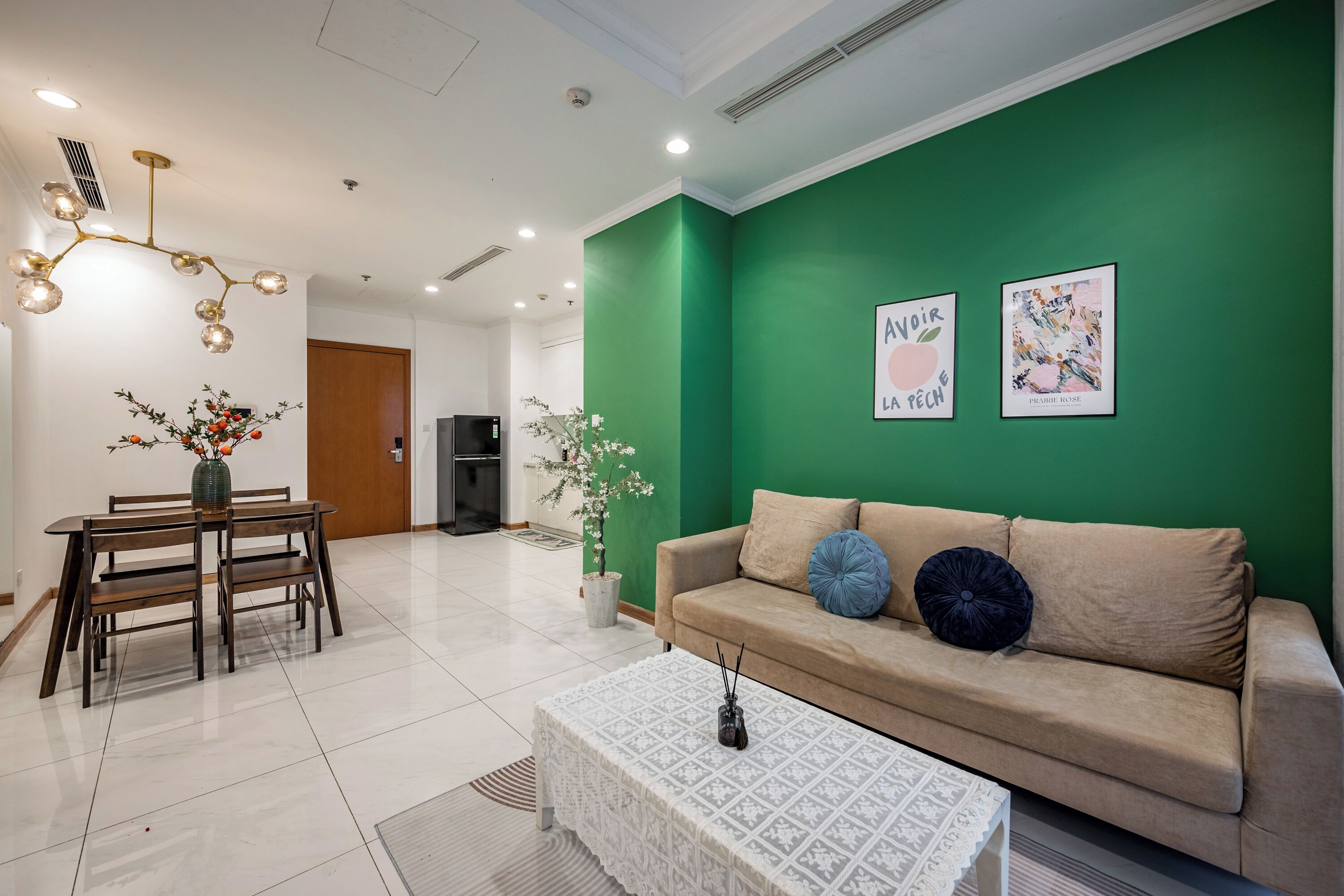 Uma sala de estar com uma parede verde, um sofá bege, uma mesa de centro branca e uma área de jantar com uma mesa de madeira e bancos.