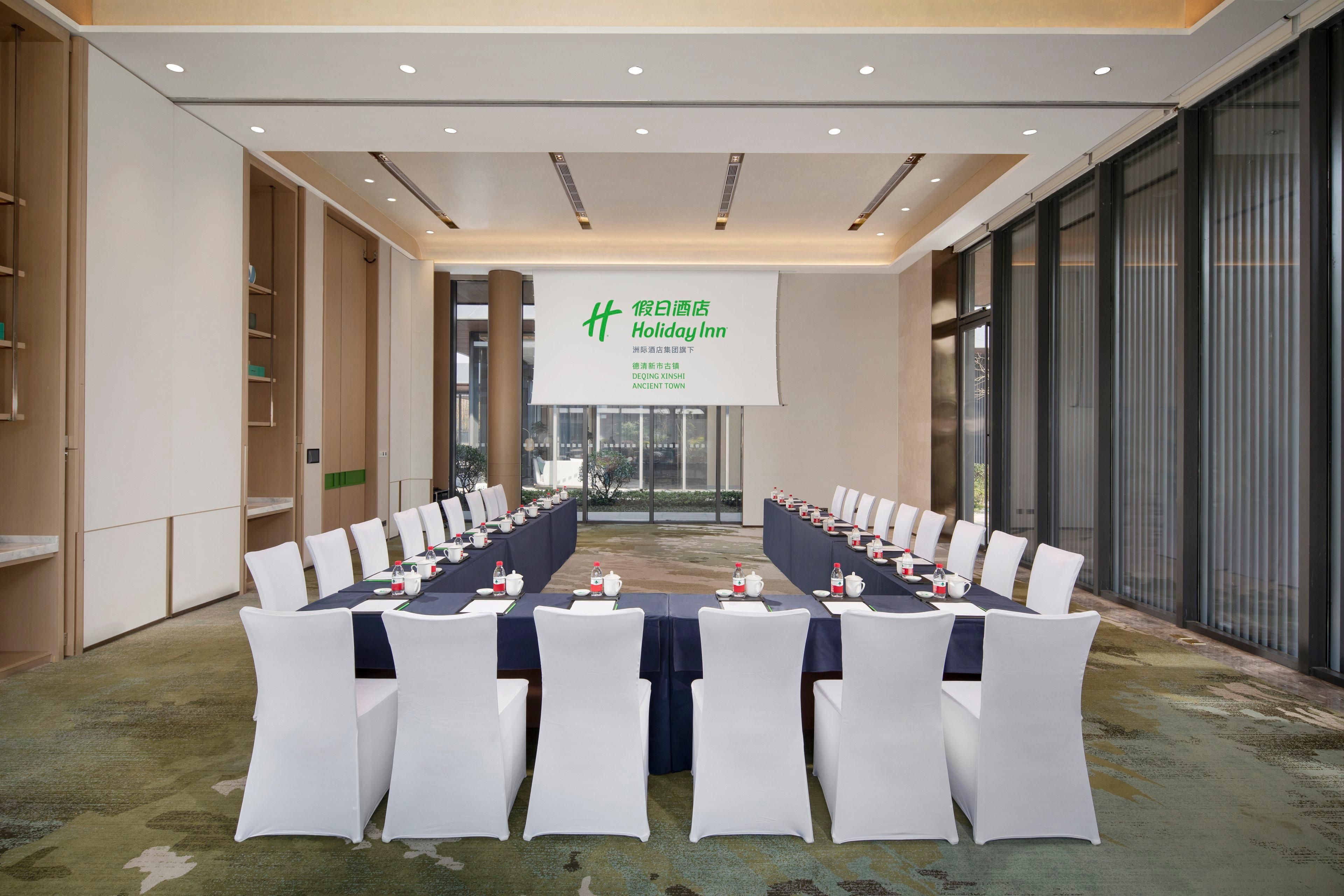 Uma sala de conferências com uma mesa comprida preparada para uma reunião, cadeiras brancas e um banner com o logo Holiday Inn.