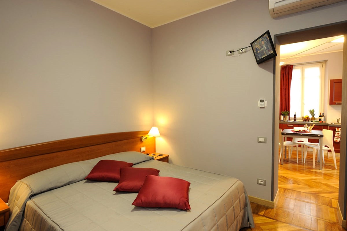 Quarto de hotel com cama, mesa de cabeceira com abajur, cabeceira de madeira e vista para a cozinha e sala de jantar através de uma porta aberta.