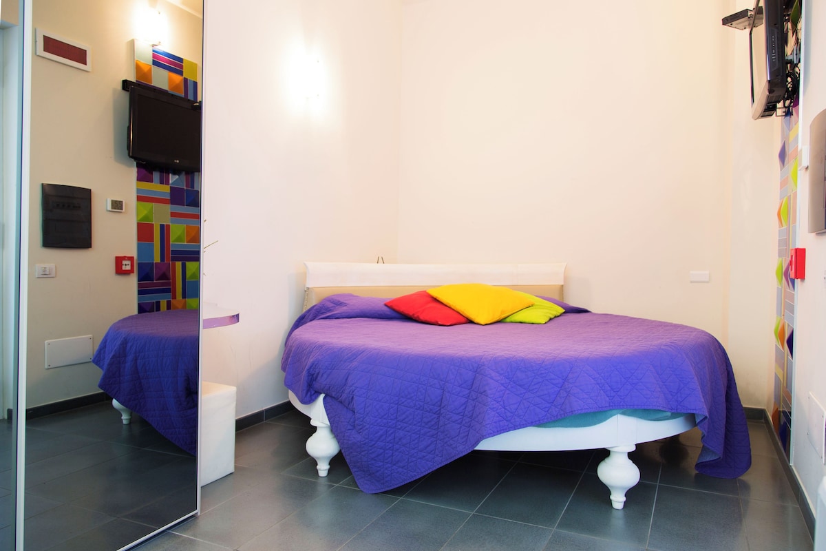 Quarto com cama, um pequeno banquinho branco, TV fixada na parede e decoração colorida com padrão geométrico.