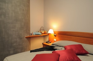 Quarto de hotel com cama, mesa de cabeceira, luminária e prateleira com objetos pessoais.