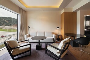 Quarto de hotel moderno com sofá, duas poltronas, mesa de centro e mesa de jantar. Há uma janela grande com vista para uma paisagem verde e uma estrada.