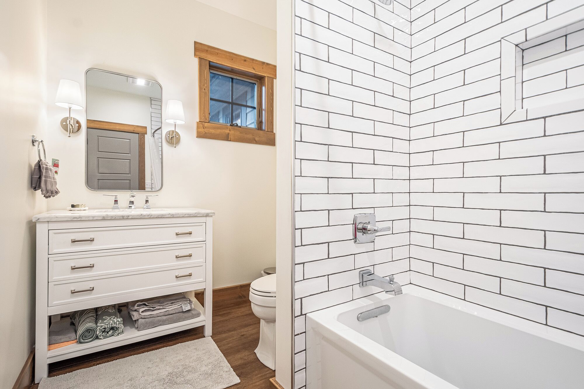 Uma casa de banho com toucador branco, espelho, janela e área de duche com azulejos brancos.
