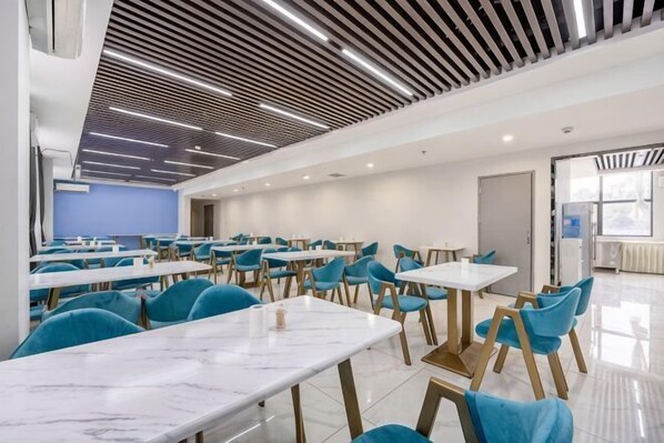 Uma área de refeições moderna com mesas de mármore e cadeiras azuis, teto curvo com iluminação linear e interior claro e impecável.