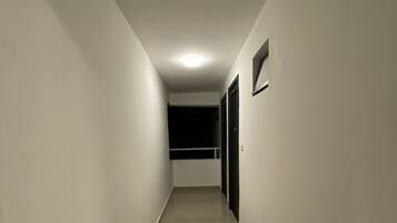 Um corredor estreito e bem iluminado com piso refletivo, conduzindo a um cômodo escuro com uma porta no final.