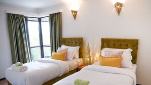 Quarto de hotel com duas camas, cada uma com lençóis brancos e uma toalha verde. Apresenta luminárias decorativas na parede e uma janela com cortinas.