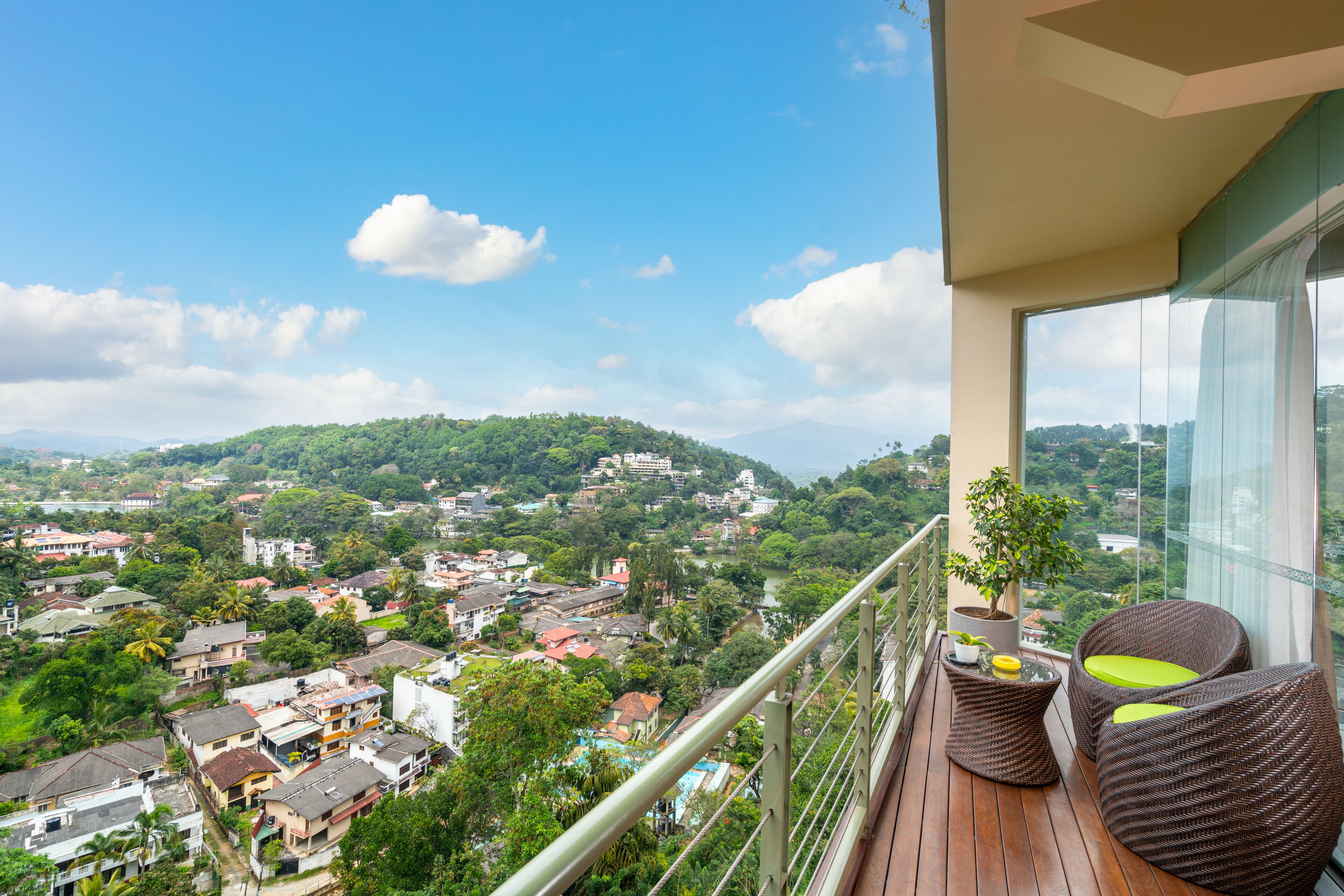 Uma varanda com móveis de vime, um vaso de planta e vista para uma cidade com edifícios e vegetação.