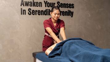 Sessão de massagem com uma pessoa deitada em uma mesa de tratamento coberta por uma toalha azul, e uma parede com a inscrição 'Awaken Your Senses In Serenity'.