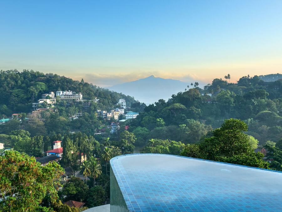Vista de uma paisagem montanhosa com um edifício grande em primeiro plano, cercado por vegetação densa e céu claro.