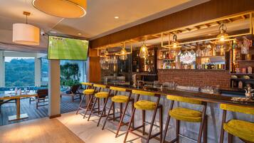 Uma área de bar com banquetas de madeira, uma tela de TV exibindo um jogo esportivo e vista para o exterior.