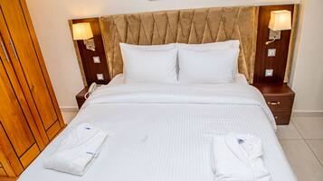 Cama bem arrumada com lençóis brancos e duas toalhas dobradas sobre ela. Há duas mesas de cabeceira com abajures, um guarda-roupa de madeira e um quadro na parede.