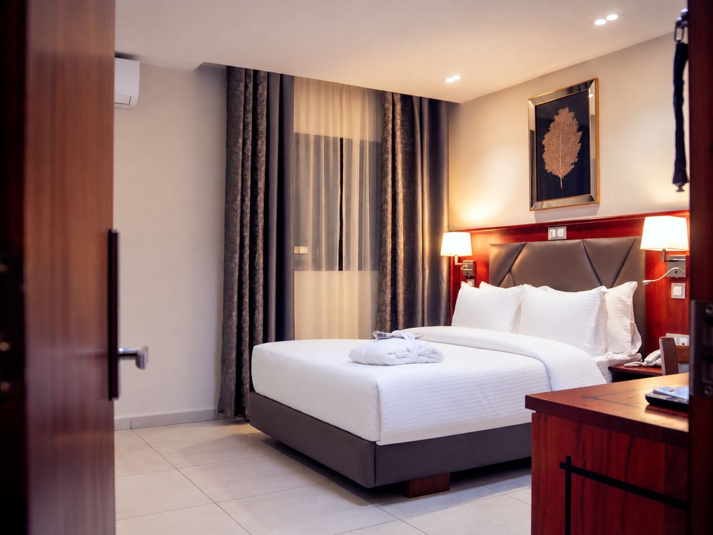 Quarto de hotel com cama, abajures, mesa de cabeceira de madeira e uma obra de arte emoldurada na parede.