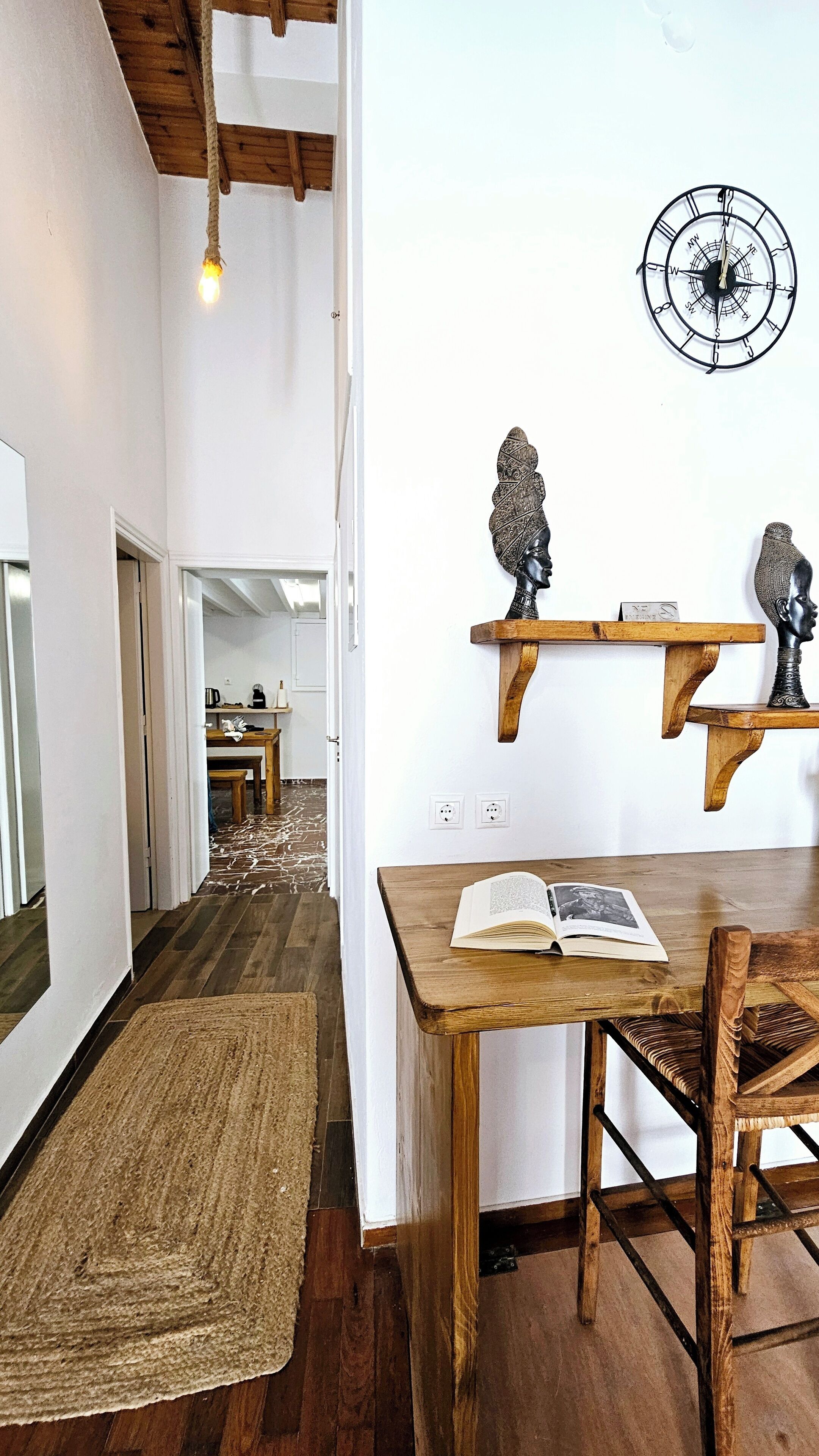 Uma mesa de jantar de madeira com um livro e uma escultura decorativa de cabeça, um relógio de parede e um candeeiro pendente num quarto com soalho de madeira e parede branca.