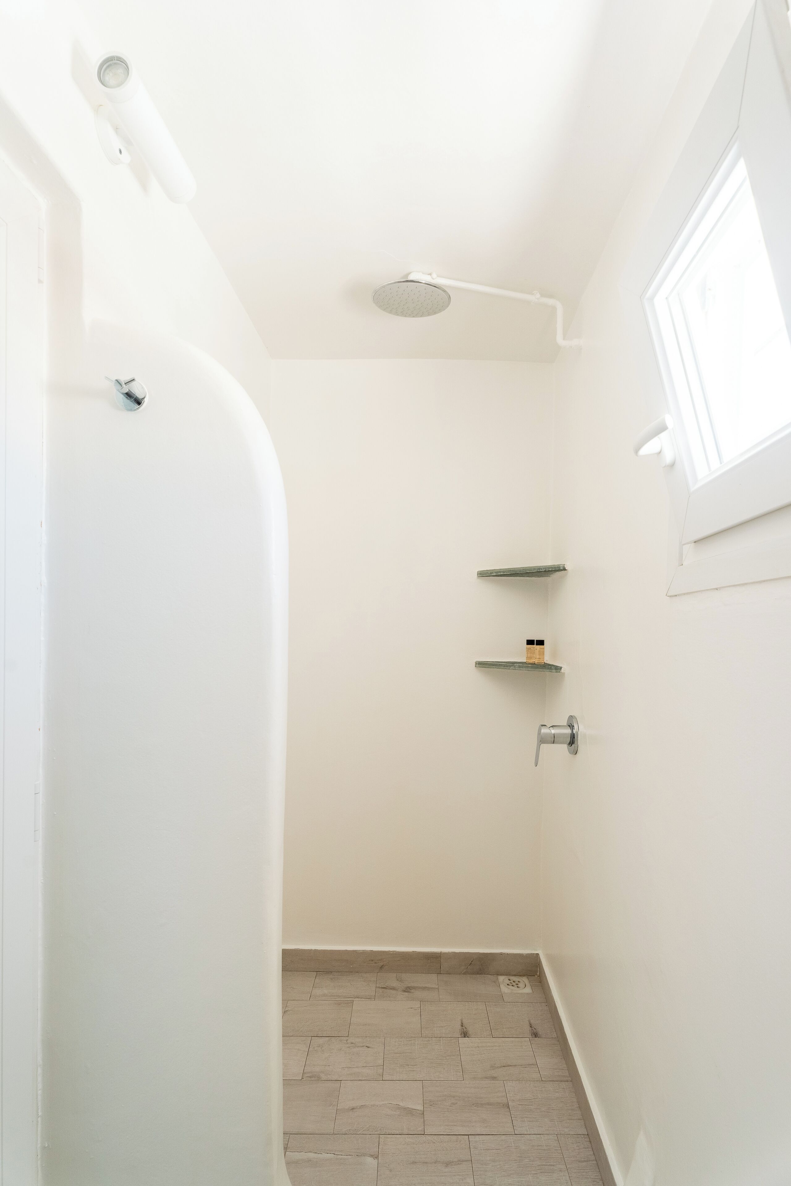 Um banheiro pequeno e branco, com chuveiro, janela e piso revestido de azulejos.