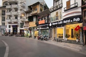 Vista da rua com um hotel chamado 'Queen Cafe Hotel & Pub', apresentando uma fachada com uma grande janela de vidro e uma decoração com uma bandeira vermelha.