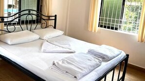 Uma cama de solteiro com estrutura de metal, roupa de cama branca e duas janelas com cortinas.