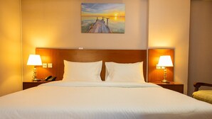 Quarto de hotel com uma cama grande, duas mesas de cabeceira com luminárias, uma pintura de um pôr do sol sobre um cais e uma cabeceira de madeira.
