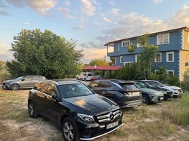 Um estacionamento com vários carros, incluindo um SUV preto Mercedes, e um edifício de dois andares com telhado vermelho ao fundo.
