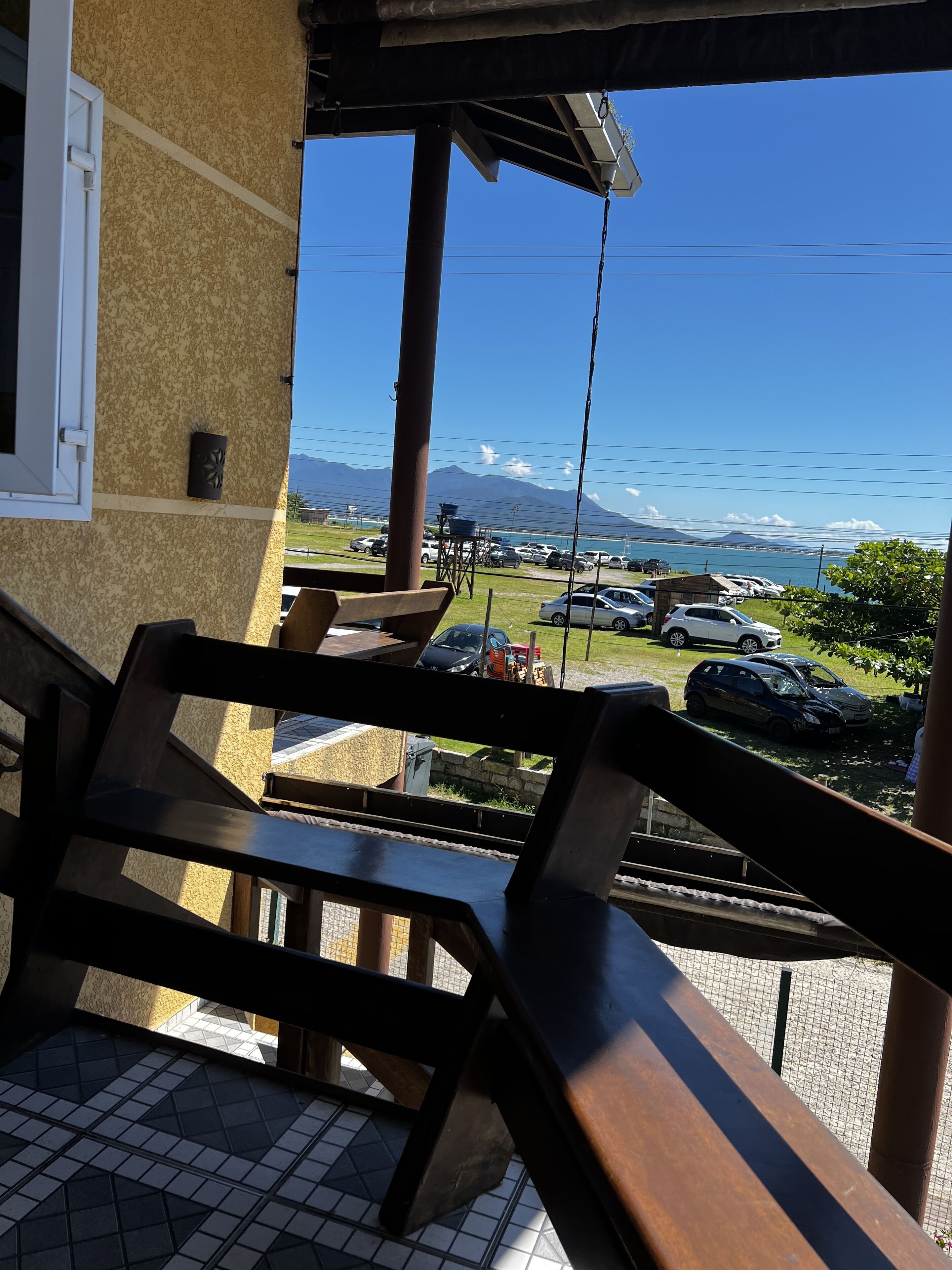 Terraço com mobiliário de madeira com vista para um estacionamento e um corpo d'água com montanhas ao longe.