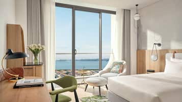 Quarto de hotel moderno com uma cama grande, uma mesa com luminária, uma cadeira e vista para o mar através de grandes janelas.