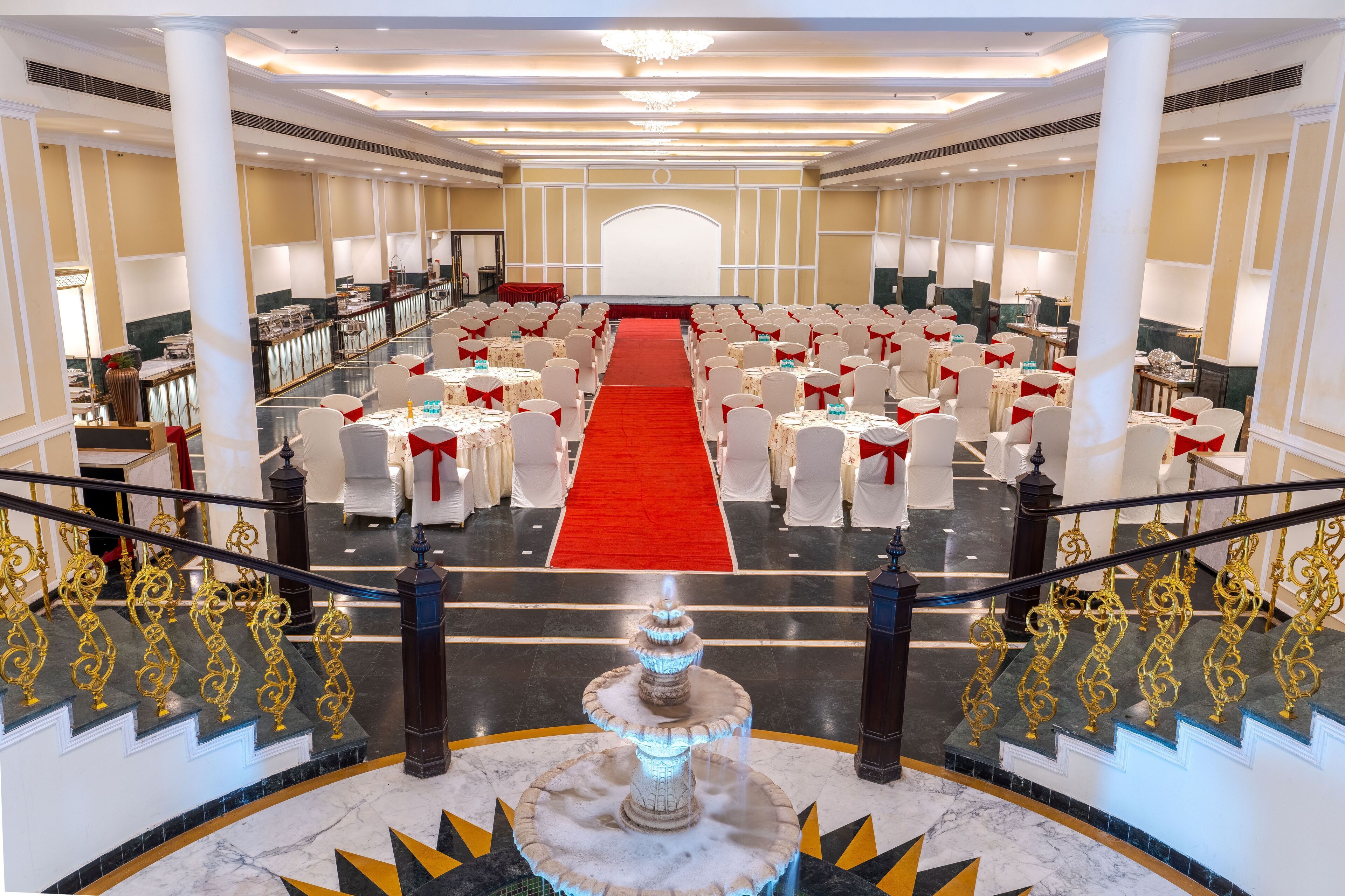 Uma luxuosa sala de baile de hotel, com tapete vermelho, cadeiras brancas e palco central.