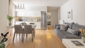 Uma sala de estar moderna com um sofá cinzento, uma mesa de jantar com cadeiras e uma cozinha ao fundo.