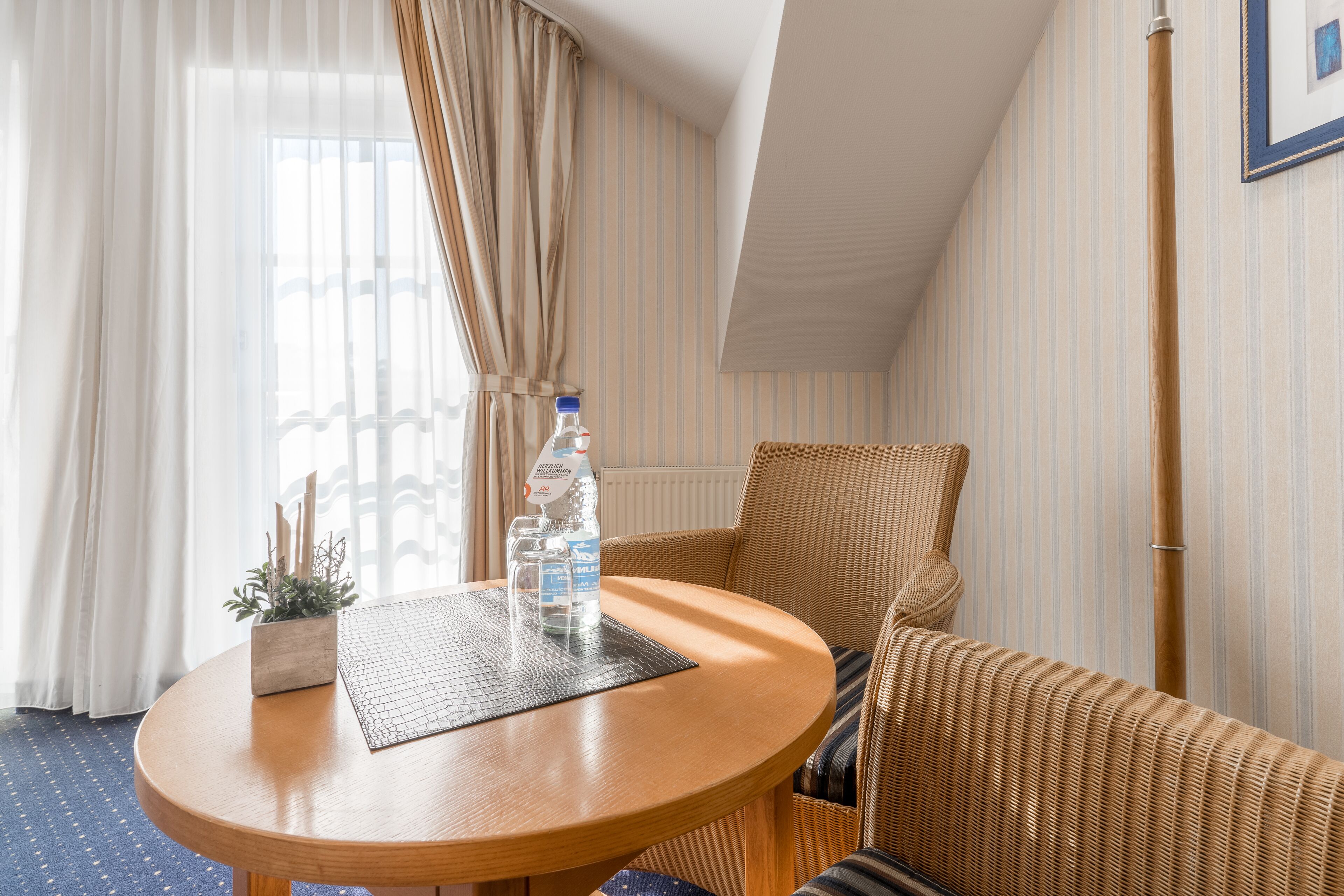 Quarto de hotel com uma mesa redonda de madeira, um sofá de vime, uma planta em vaso e uma garrafa de água.